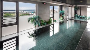 Amplo espaço interno com grandes janelas que oferecem vista para o mar e edifícios, piso lustroso e plantas em vasos ao longo das paredes.