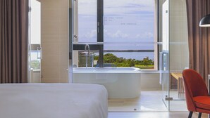 Quarto de hotel moderno com uma cama grande, banheiro com paredes de vidro e banheira, e vista para o mar.
