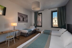 Quarto de hotel com duas camas, uma escrivaninha, uma cadeira, um abajur e vista para um edifício pela janela.