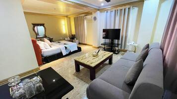 Quarto de hotel com sala de estar, sofá, mesa de centro, televisão e cama com mesa de cabeceira.
