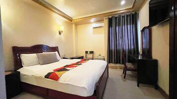 Quarto de hotel com cama grande, mesas de cabeceira, escrivaninha, cadeira, televisão e cortina.