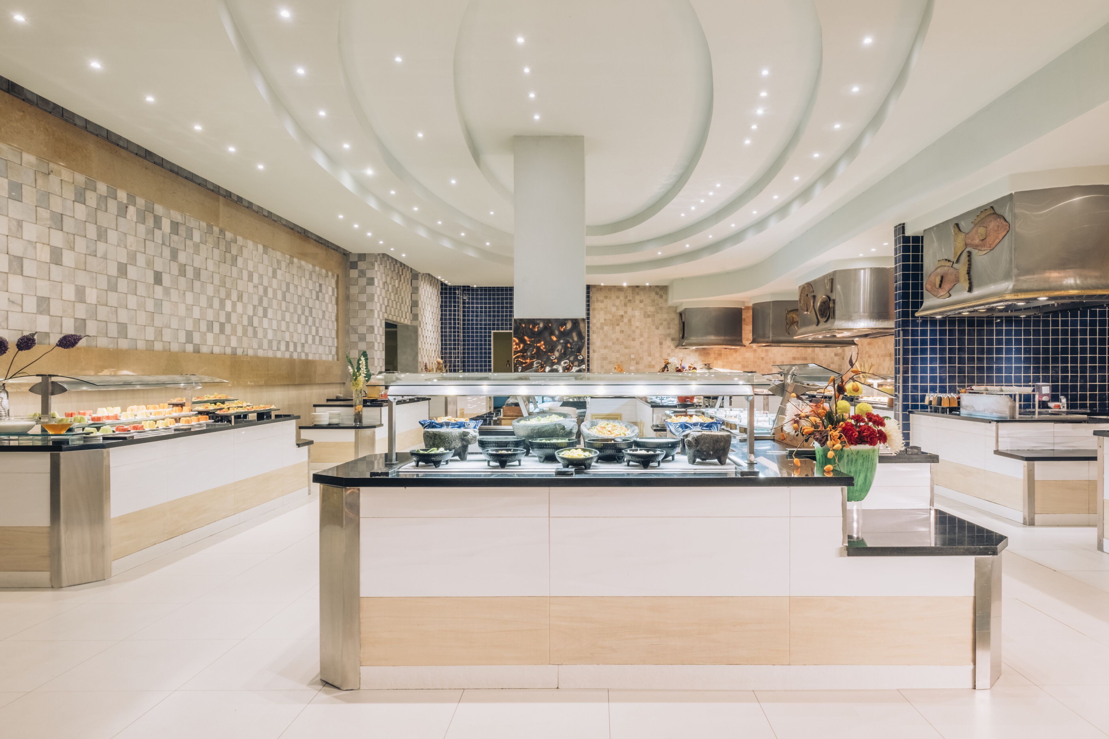 Uma área de buffet moderna com uma ilha central exibindo diversos alimentos, rodeada por uma parede revestida de azulejos e um teto decorativo.