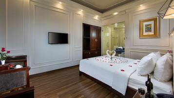 Quarto de hotel com chão de madeira, uma cama com colcha branca e decorações em formato de coração, uma TV fixada na parede e um espelho com estrutura de vidro.