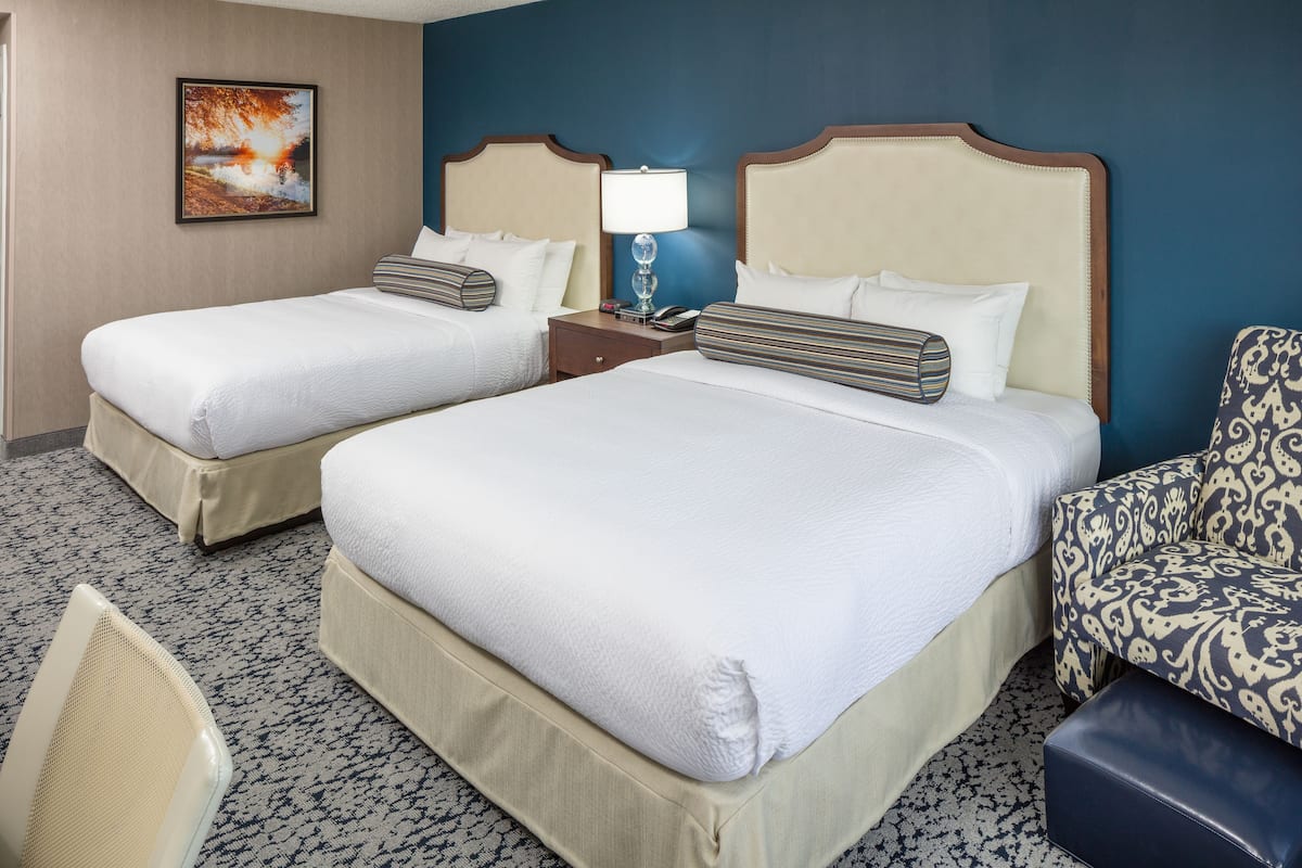 Quarto de hotel com duas camas, uma mesa de cabeceira com abajur, uma cadeira e uma poltrona estampada.
