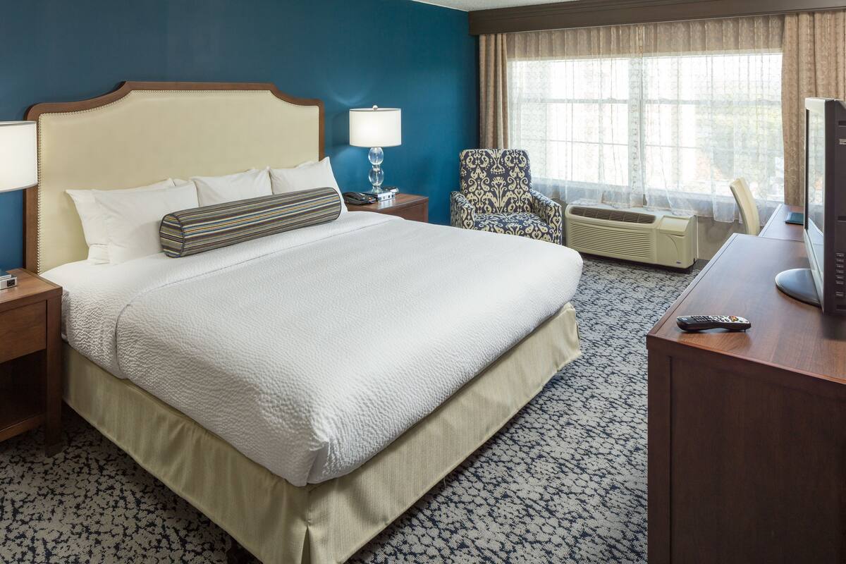 Quarto de hotel com uma cama grande, uma escrivaninha com computador, uma cadeira e uma janela com cortinas.