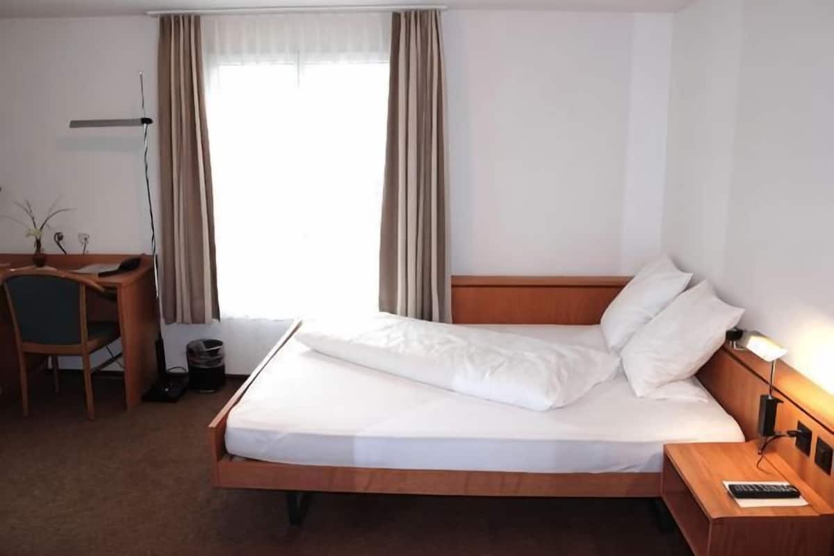 Quarto de hotel com cama, escrivaninha, cadeira, abajur e janela com cortinas.