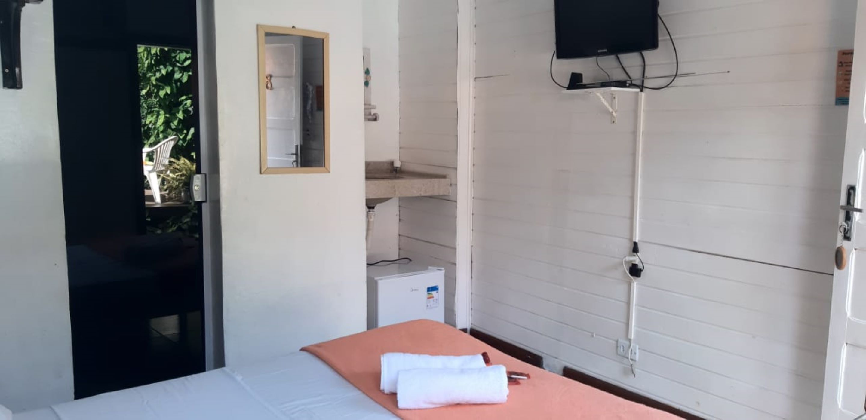 Quarto com cama, uma mesinha com toalha, TV fixada na parede e porta que dá para uma varanda.