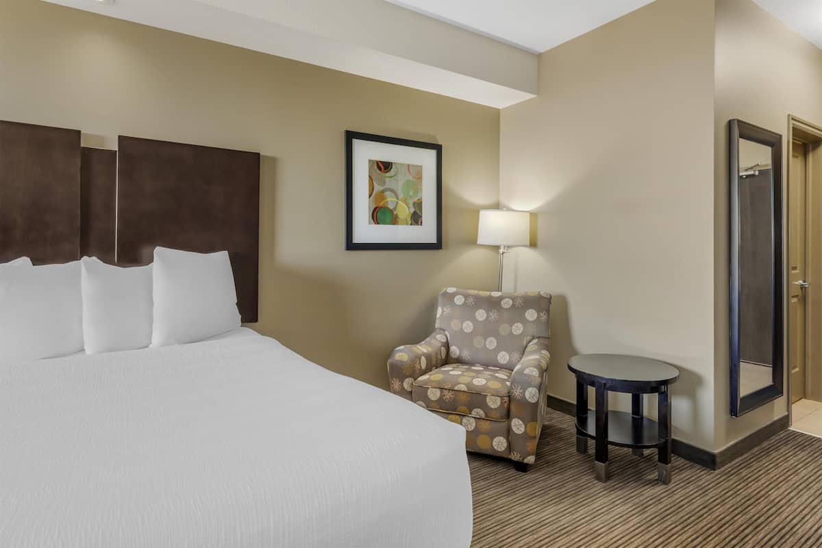 Quarto de hotel com uma cama grande, uma cadeira, uma mesinha, um abajur, um quadro na parede e uma porta.