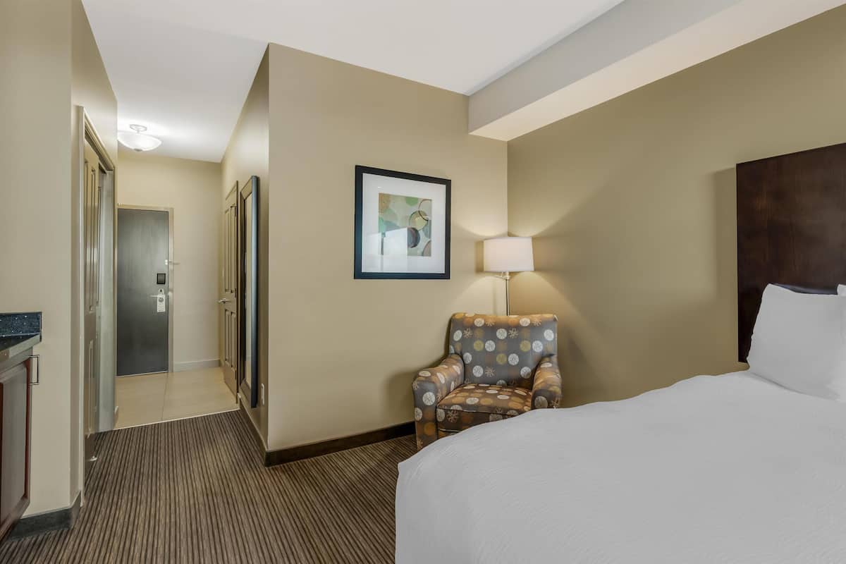 Quarto de hotel com cama, uma cadeira, luminária e porta que dá para um corredor.