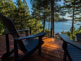 Um deck com cadeiras Adirondack com vista para um lago, árvores e terra distante.