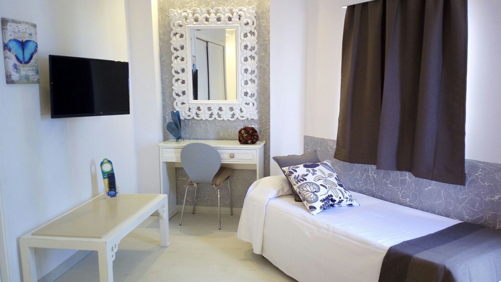 Quarto de hotel com cama, escrivaninha, cadeira, televisão e espelho.