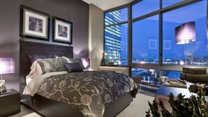Um quarto com uma cama grande, uma mesa de cabeceira, uma luminária, uma mesa com vista para a cidade e quadros nas paredes.