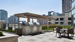 Um terraço no telhado com churrasqueira e área de estar, rodeado por edifícios altos.