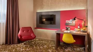 Quarto de hotel com televisão presa à parede, escrivaninha com cadeira, uma cadeira vermelha, uma cadeira amarela e um chão estampado.