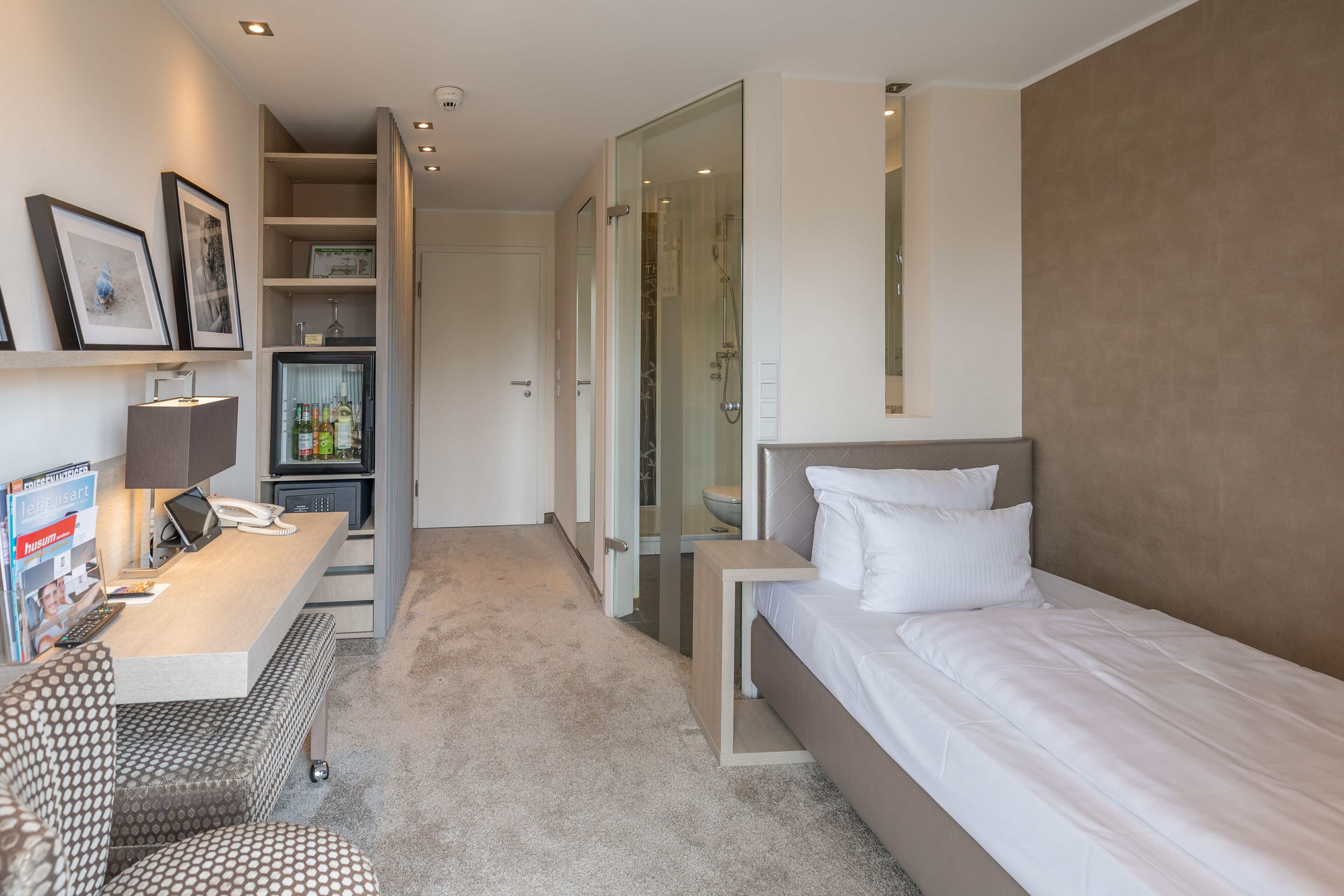 Quarto de hotel moderno com cama, mesa de cabeceira, escrivaninha com abajur, cadeira e box de vidro para o chuveiro.