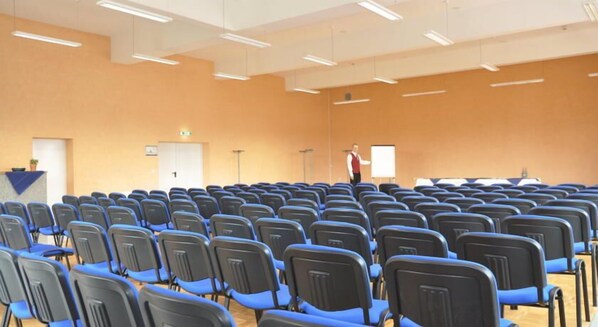 Uma sala de conferências com fileiras de cadeiras azuis, um púlpito e um quadro branco.