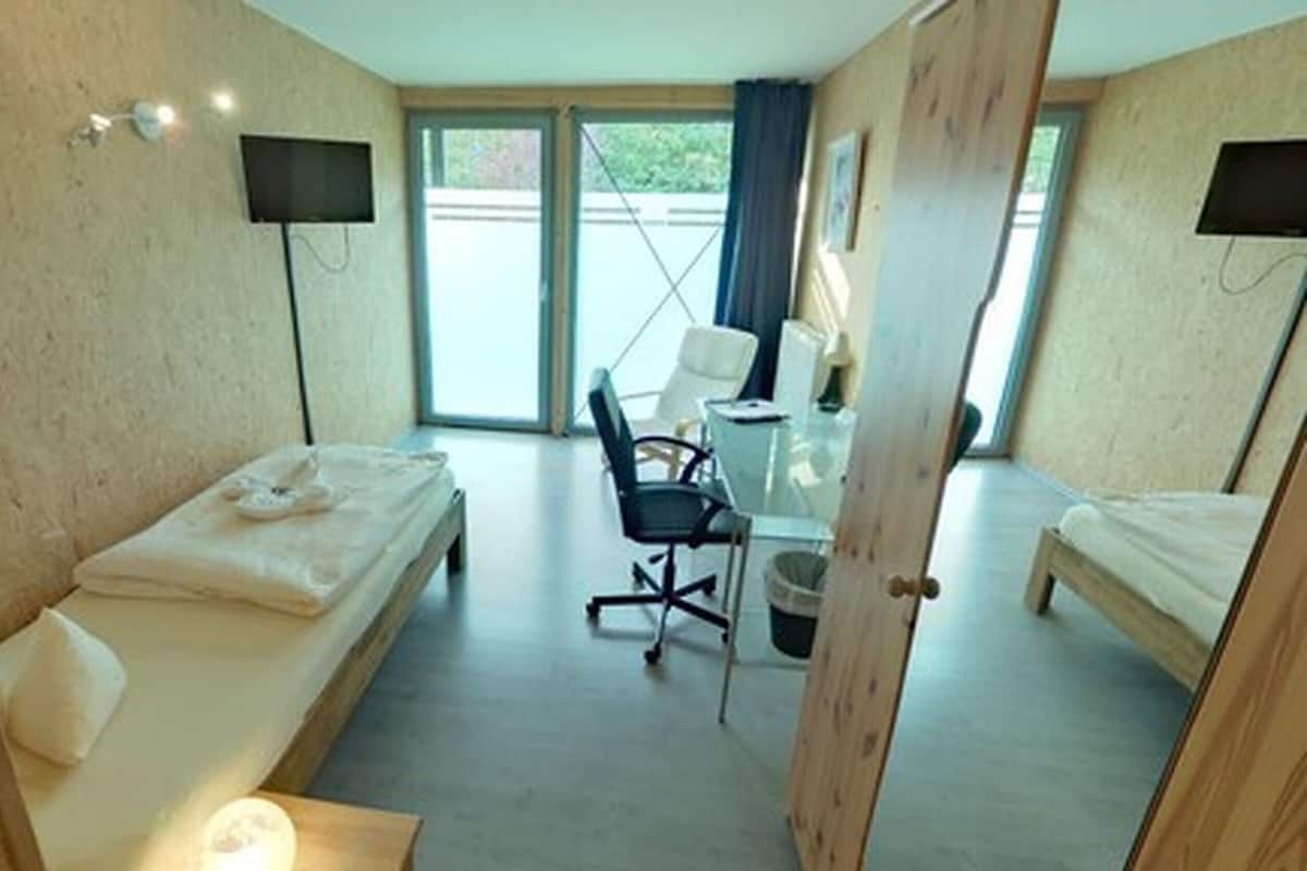 Quarto com duas camas, uma escrivaninha, uma cadeira e televisão na parede.