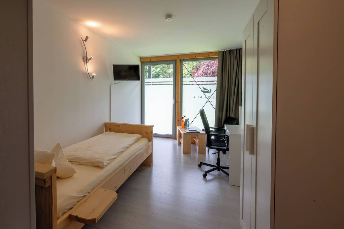 Quarto individual com uma cama, escrivaninha, cadeira e televisão fixada na parede.