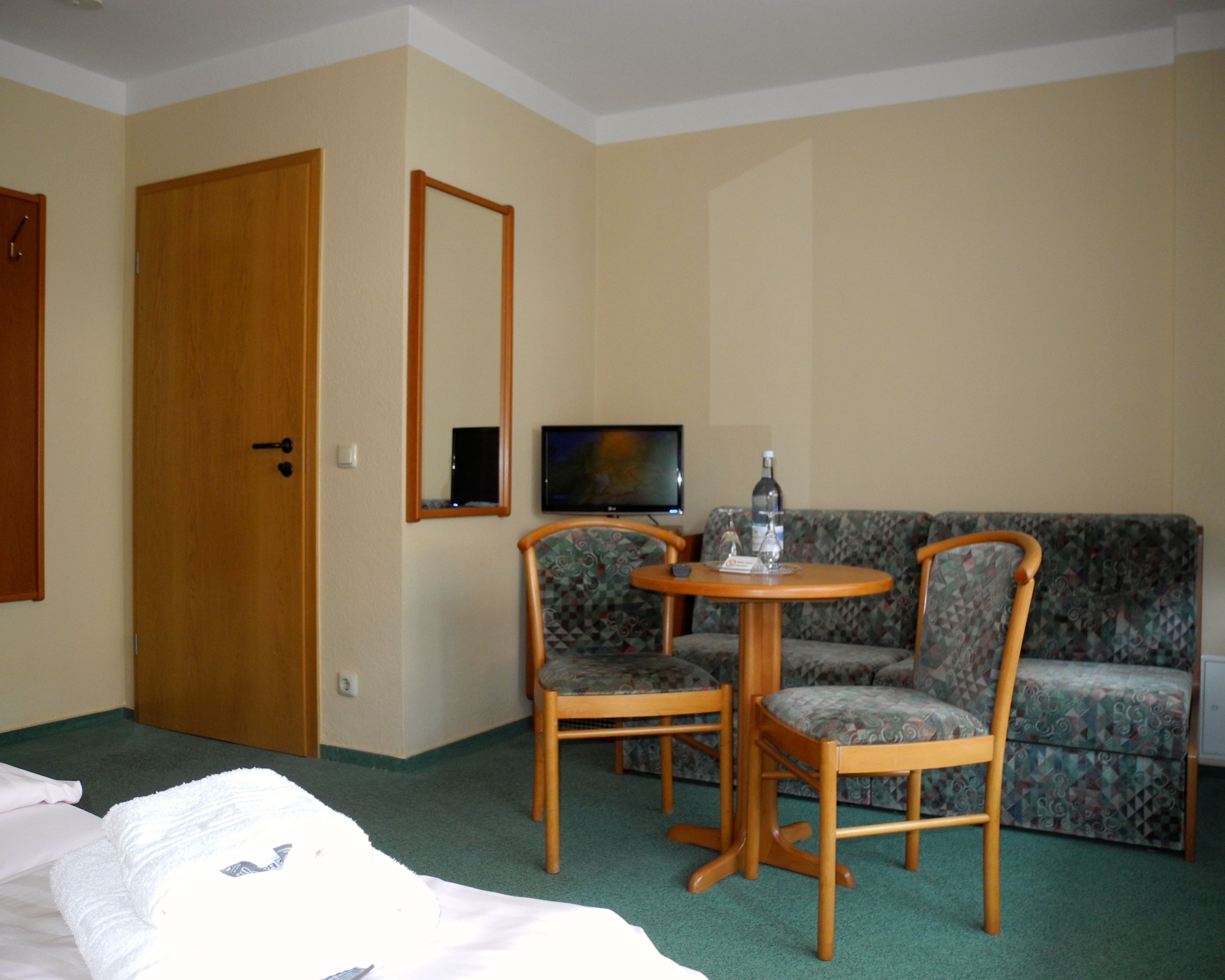 Quarto de hotel com uma cama, televisão, sofá, duas cadeiras e uma mesa pequena com uma garrafa e copos.