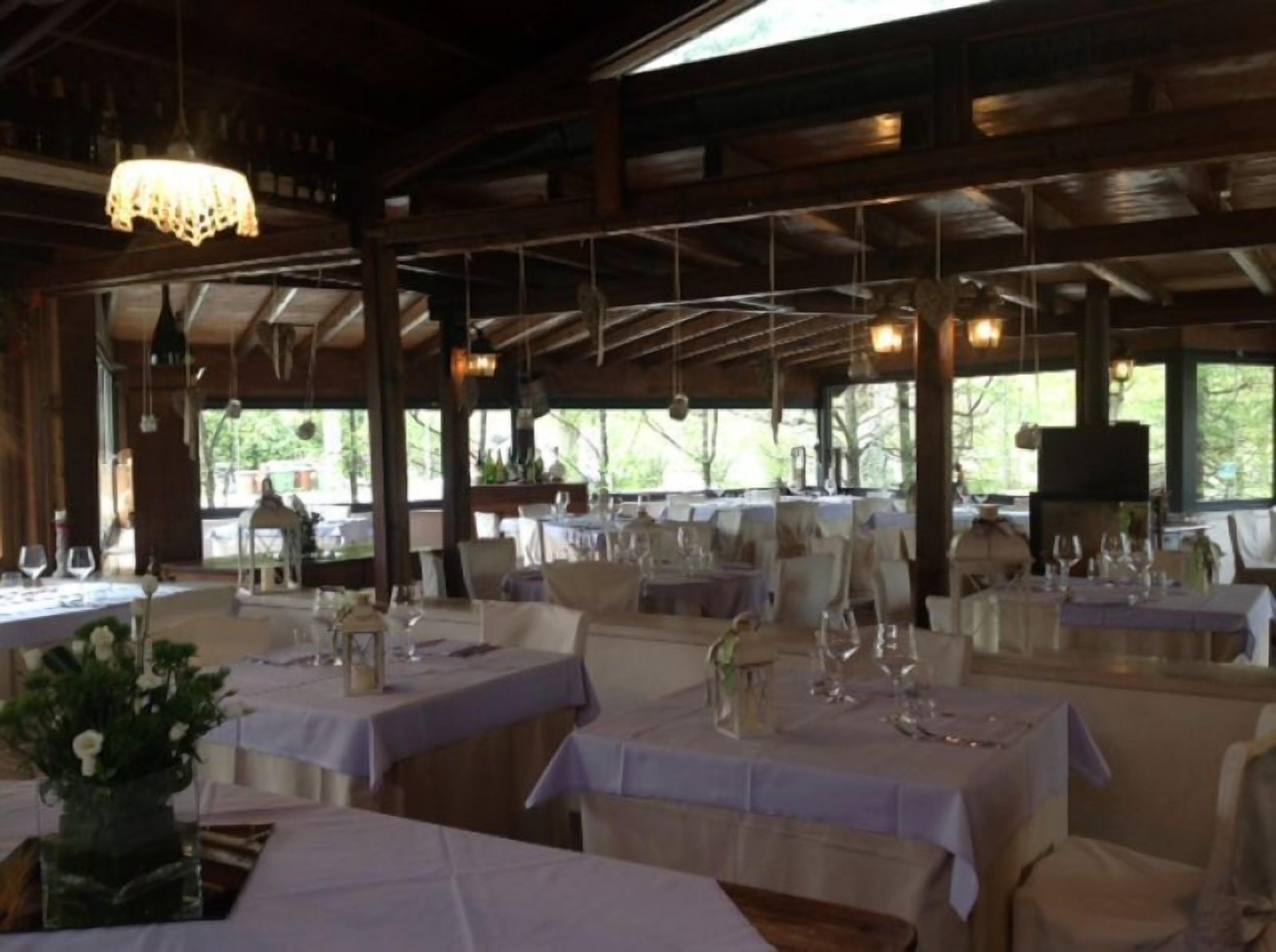 Um espaço de refeições interno com mesas preparadas para o serviço, grandes janelas e luminárias pendentes.