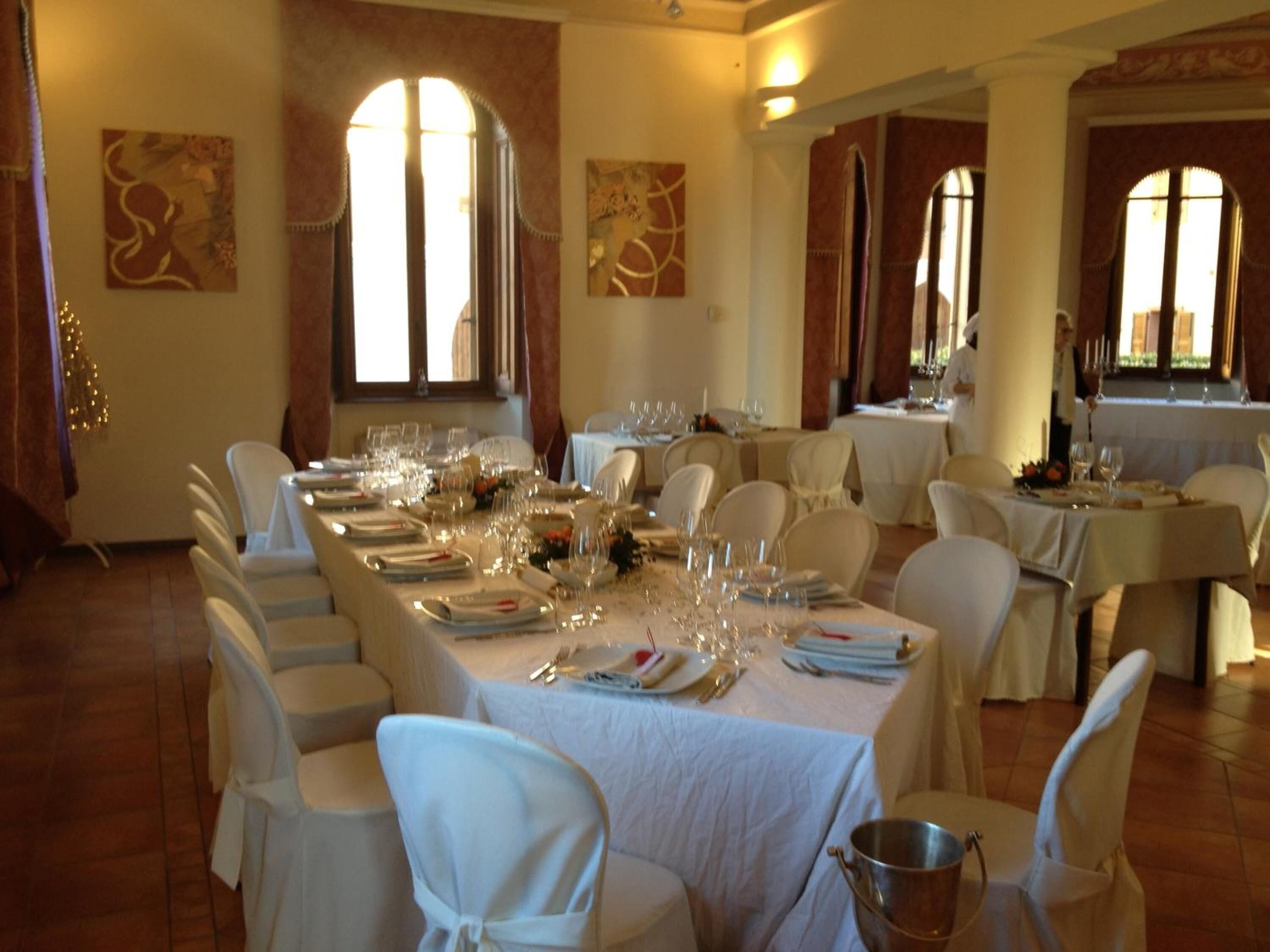 Uma sala de jantar formal com várias mesas postas para refeição, cadeiras brancas e grandes janelas que permitem entrada de luz natural.