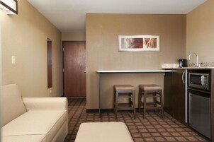 Quarto de hotel compacto com cozinha compacta, sofá, dois bancos altos e um quadro na parede.