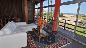 Um quarto amplo com um sofá branco, uma mesa de centro de madeira com um vaso de flores e grandes janelas com vista para o oceano e um estacionamento.