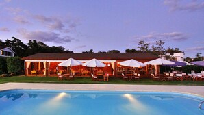 Área da piscina com mobiliário de exterior e guarda-sóis, um edifício com telhado vermelho e céu claro.