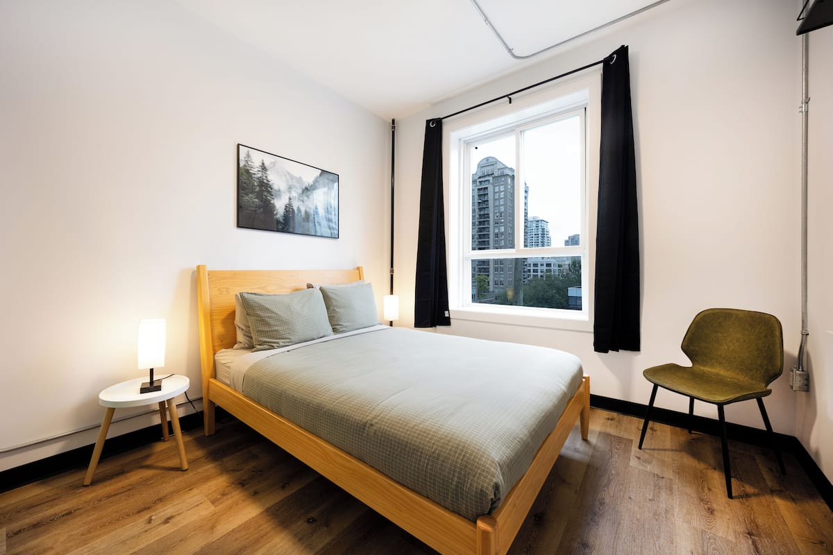 Quarto moderno com cama de madeira, mesa de cabeceira pequena, uma cadeira, janela com cortinas e vista para a cidade.
