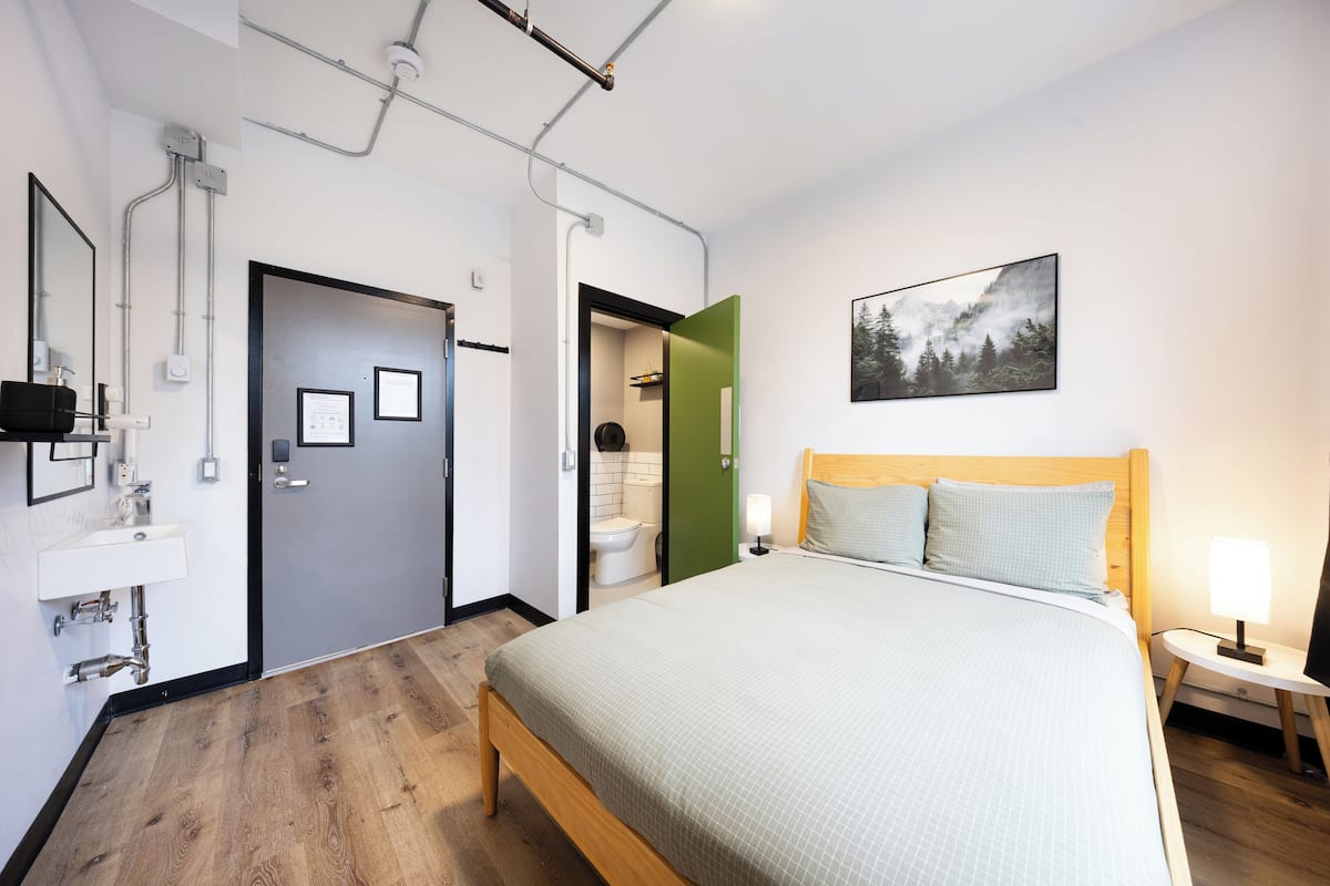 Quarto de hotel moderno com piso de madeira, uma cama grande com cabeceira, mesa de cabeceira com abajur, banheiro com vaso sanitário e pia, e um quadro na parede.