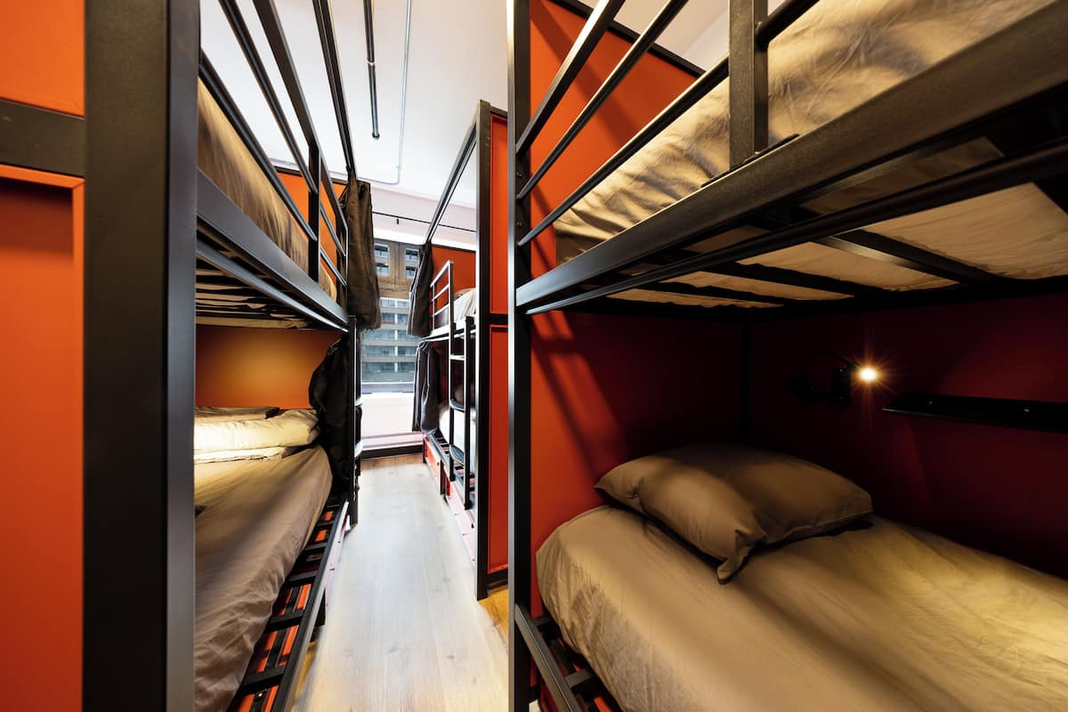 Quarto estilo dormitório com beliches, piso de madeira e vista para a cidade pela janela.