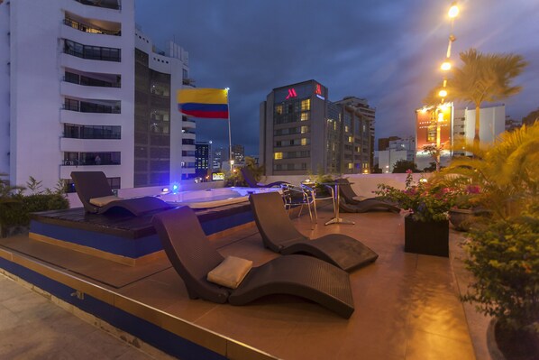 Uma área de lounge no terraço com espreguiçadeiras e uma banheira de hidromassagem, cercada por edifícios da cidade e uma bandeira.