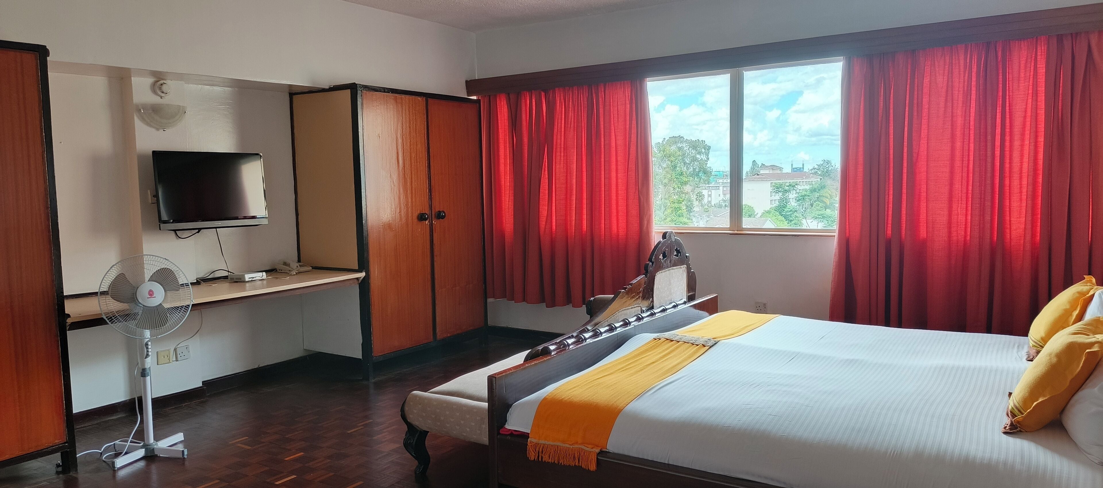 Quarto de hotel com uma cama, uma escrivaninha com televisão, um guarda-roupa e uma janela com cortinas vermelhas.