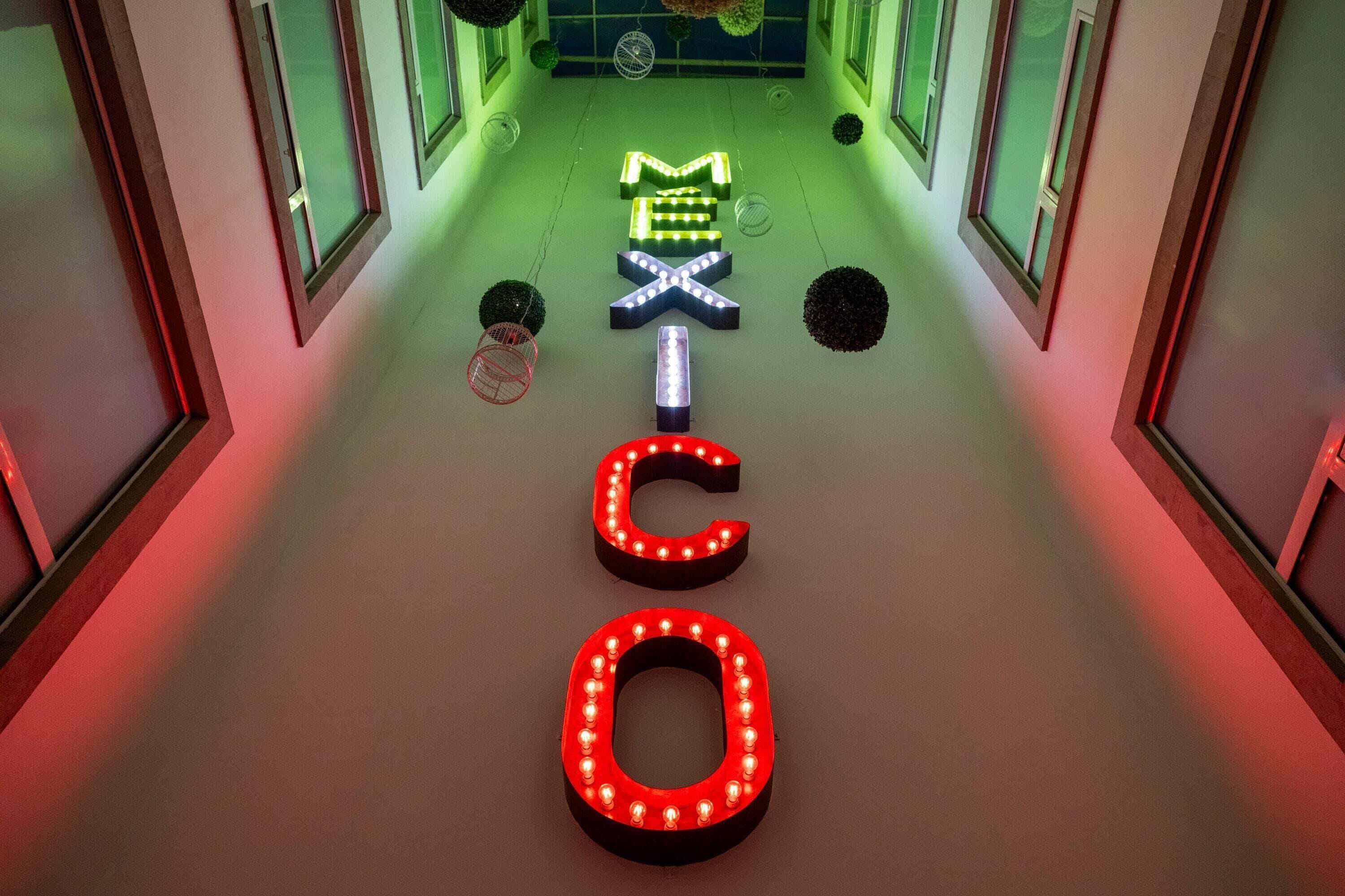 Um corredor com letras iluminadas formando 'C O' no chão, conduzindo a uma porta com um letreiro verde.