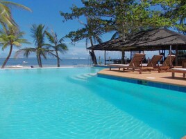 Piscina azul límpida com espreguiçadeiras e uma estrutura de palha, rodeada por palmeiras e vista para o oceano.