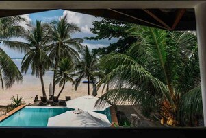 Uma área de piscina com espreguiçadeiras e guarda-sóis, com vista para uma praia com palmeiras e um mar tranquilo.