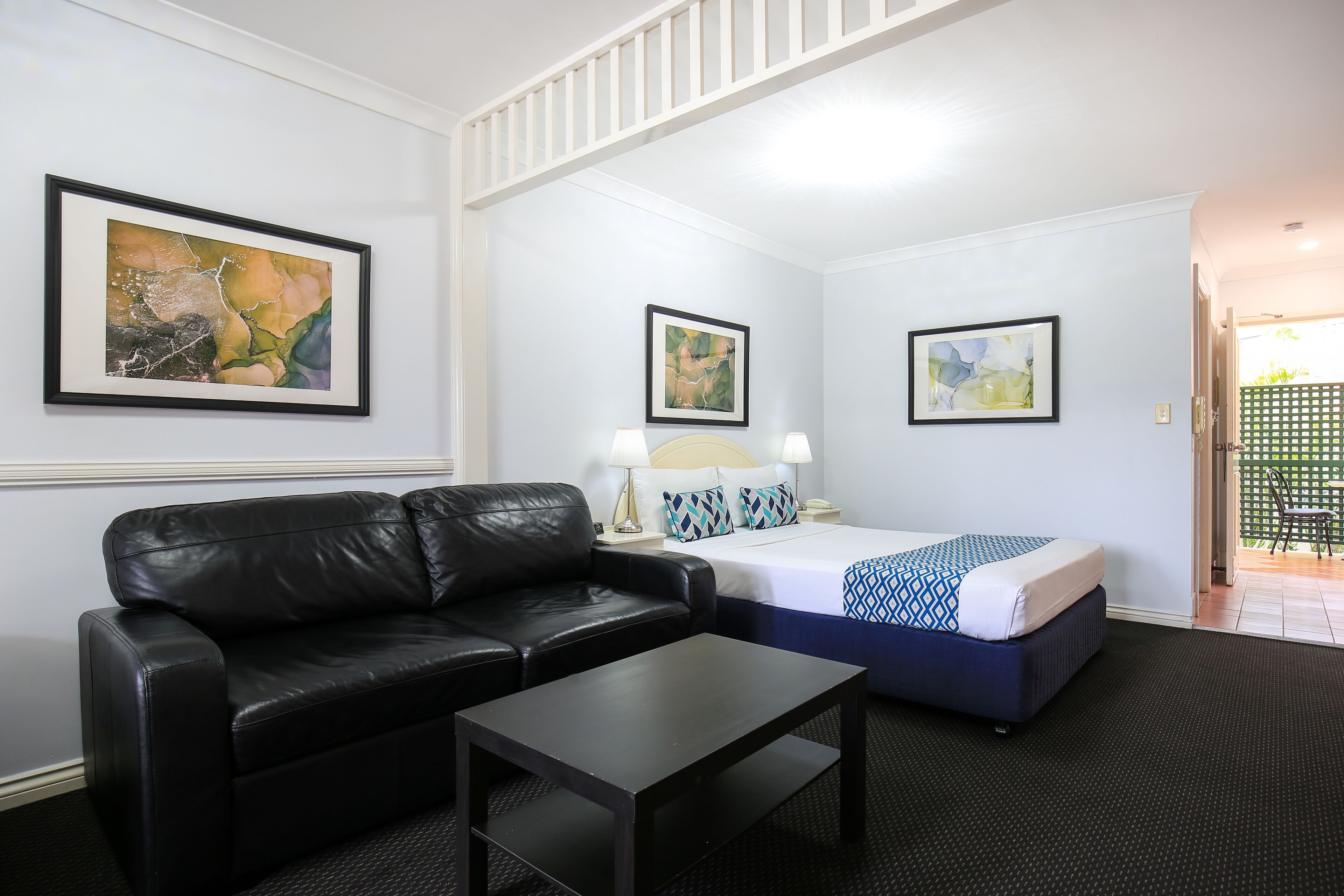 Quarto de hotel com sofá de couro preto, uma cama de solteiro com colcha de padrão azul e branco, duas pinturas abstratas emolduradas nas paredes, uma mesa pequena e vista para o exterior por uma porta de vidro.