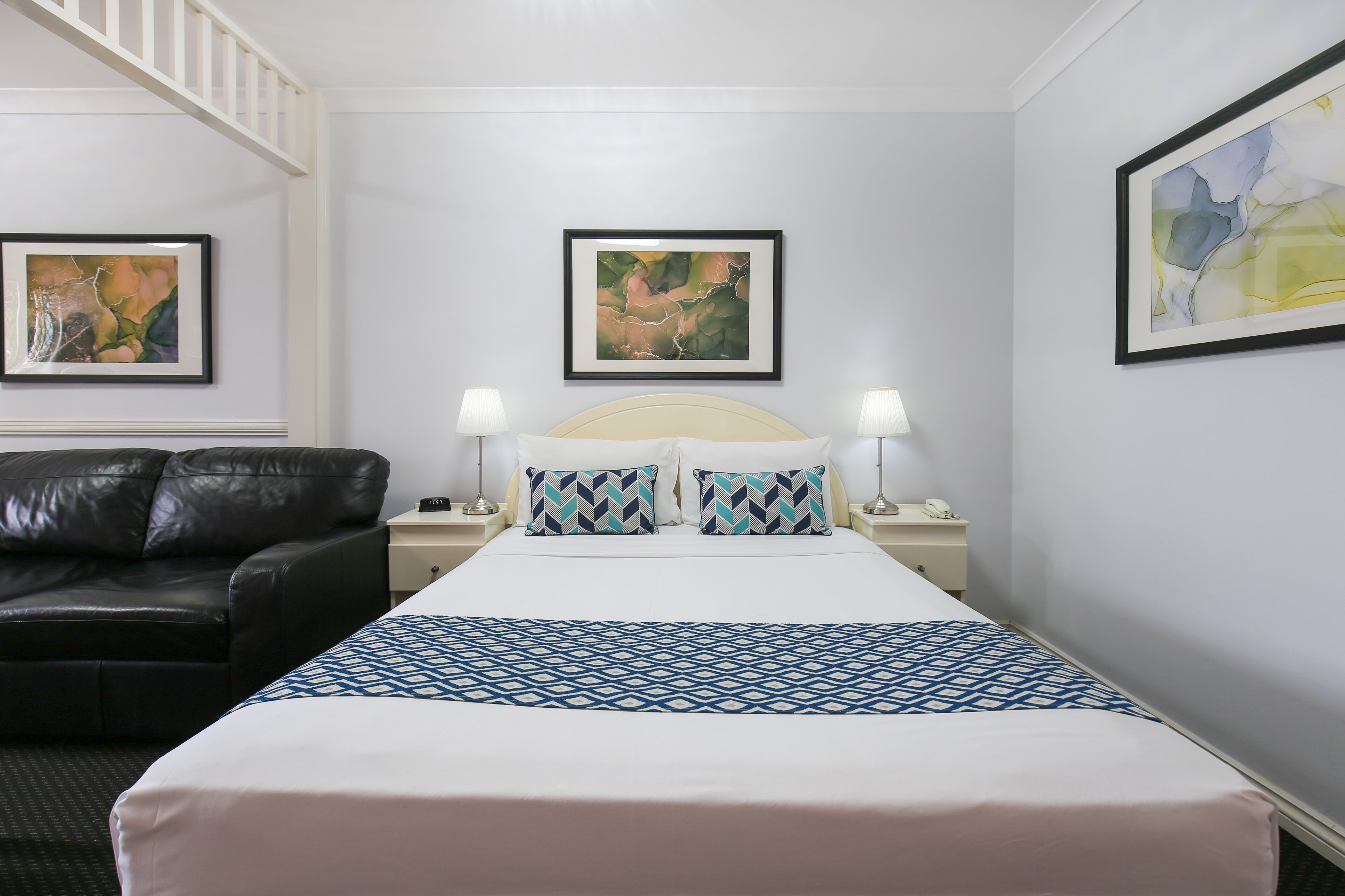 Quarto de hotel com cama, duas mesas de cabeceira com luminárias, um sofá de couro preto e três obras de arte emolduradas nas paredes.