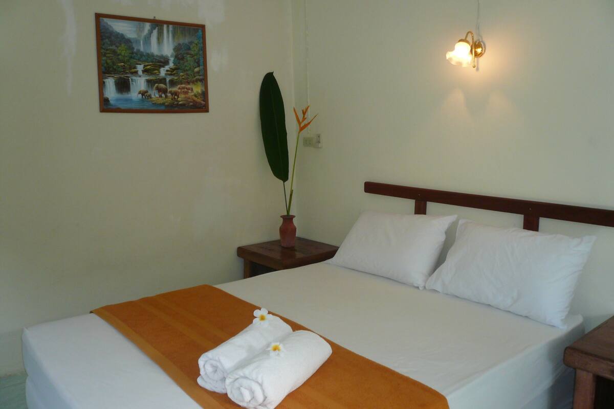Quarto de hotel com uma cama, uma pintura de uma cascata, uma mesa de cabeceira com uma planta e um candeeiro fixo na parede.