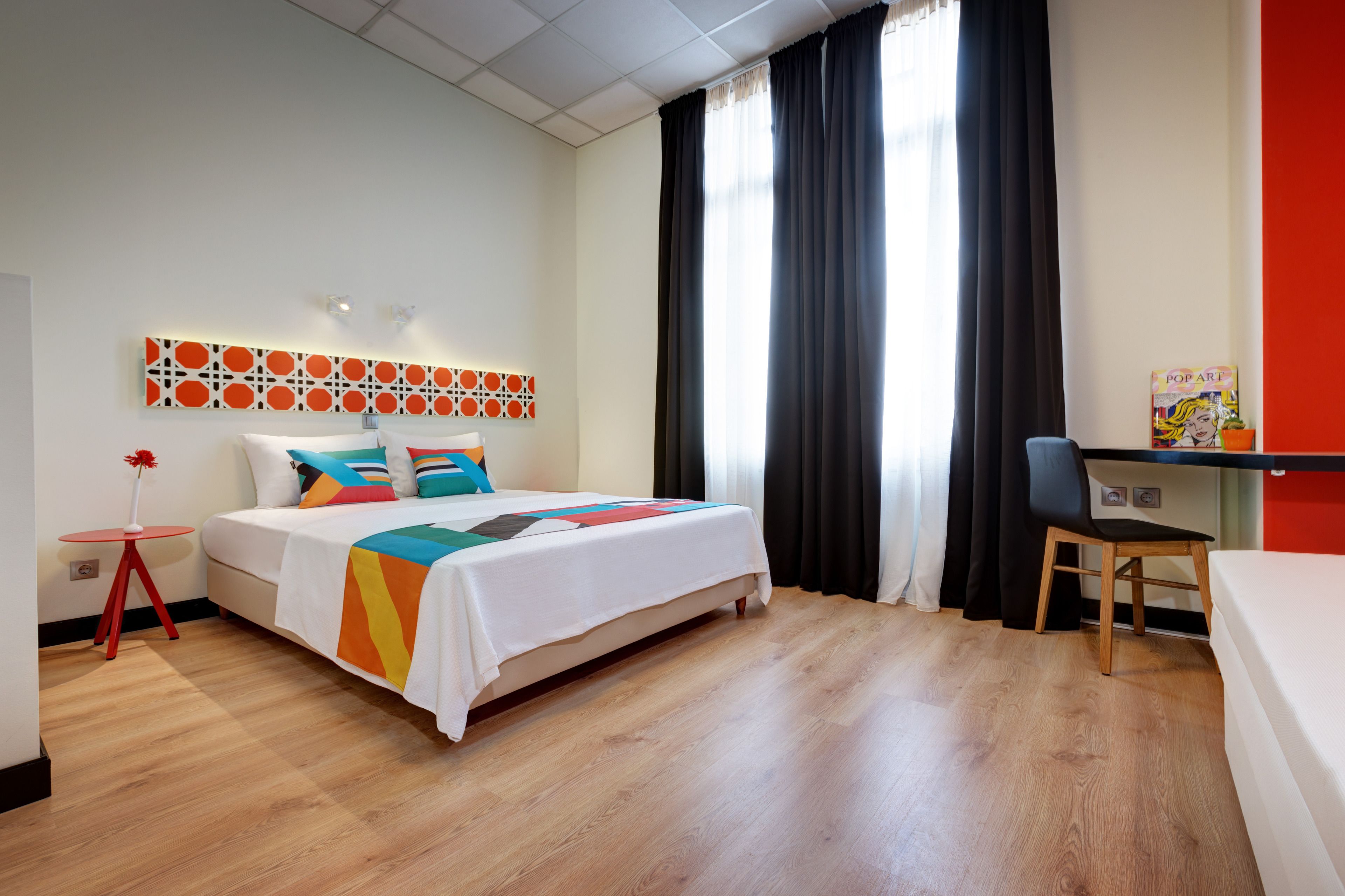 Quarto de hotel moderno com cama bem arrumada, uma secretária pequena e uma janela grande com cortinas.