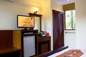 Quarto de hotel com uma cama, uma secretária com um computador, um pequeno frigorífico, uma televisão na parede e uma janela com vista para o verde.