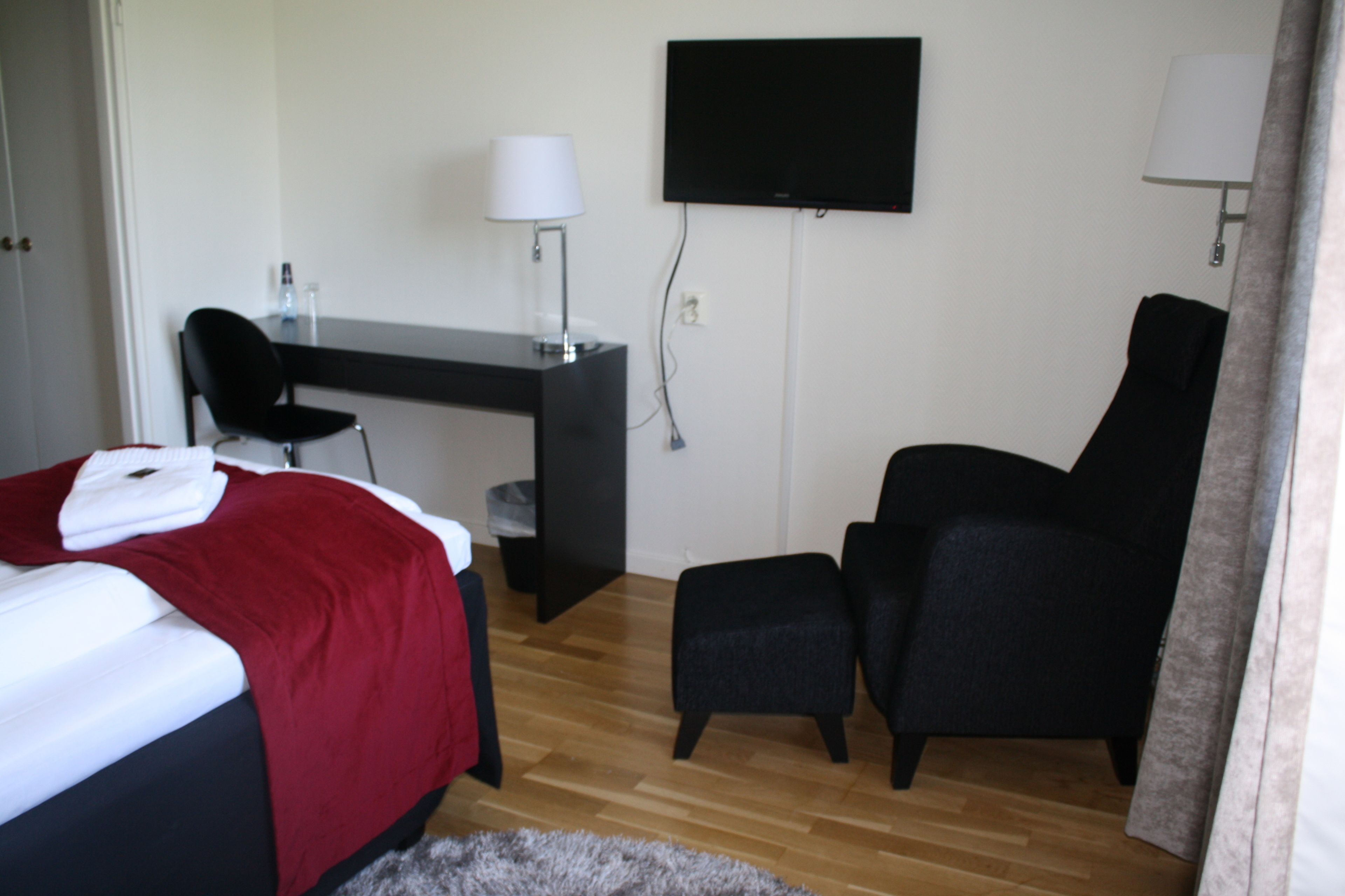 Quarto de hotel com cama, escrivaninha, cadeira, televisão e luminária.