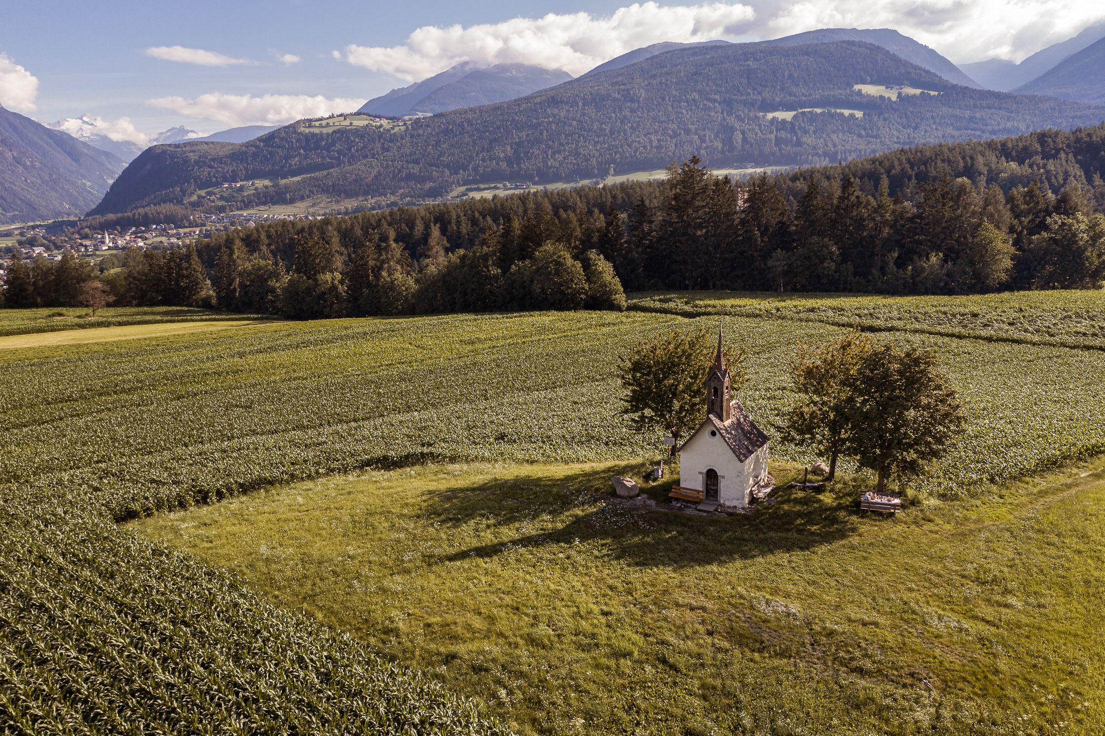 Uma pequena igreja branca com um campanário, rodeada por um campo verdejante e uma cadeia de montanhas ao fundo.
