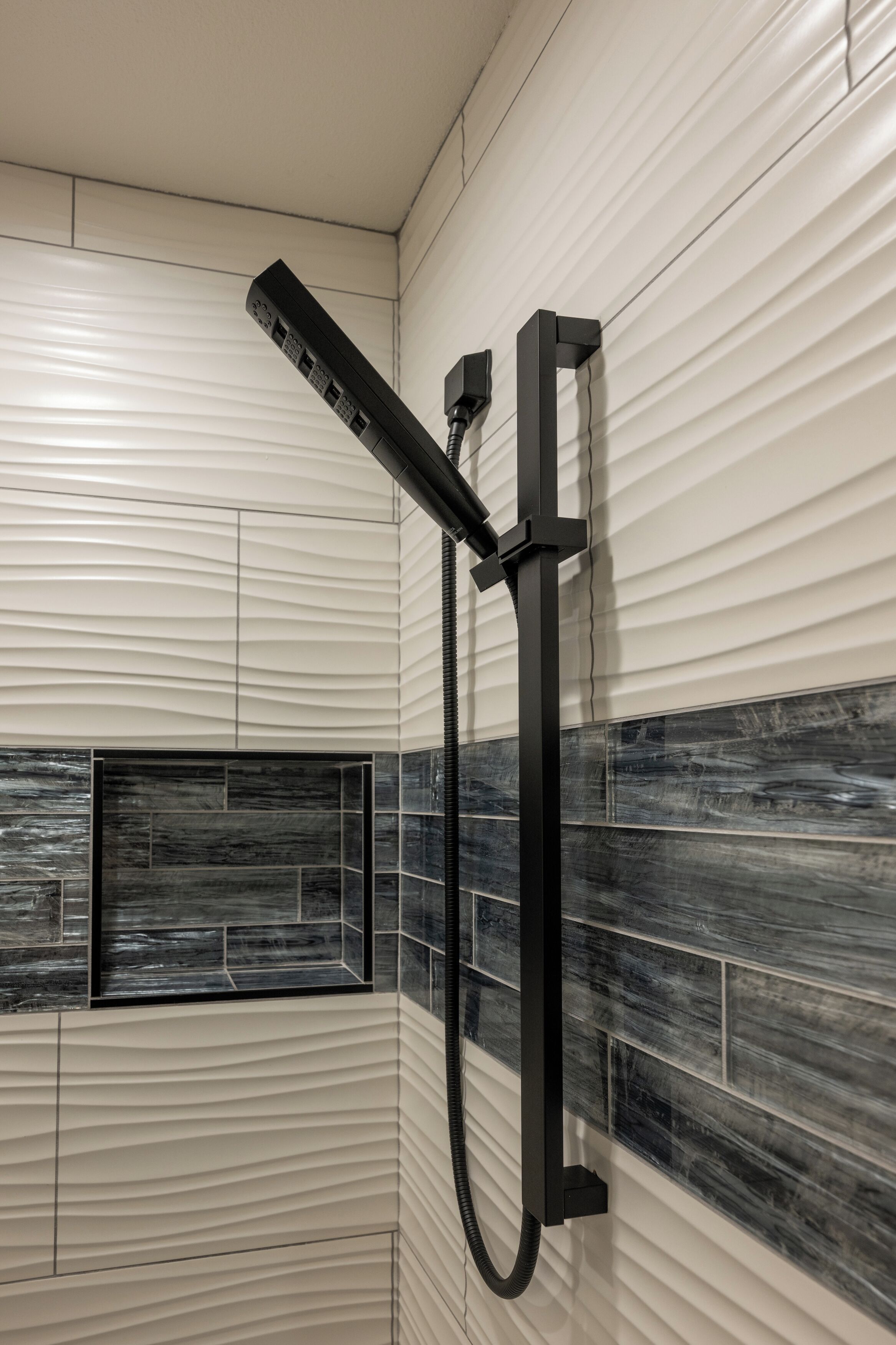 Chuveiro moderno com mangueira preta e painel de controle, em frente a uma parede com textura e uma área de azulejos escuros.