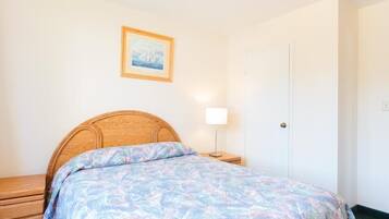 Quarto com cabeceira de madeira, cama com colcha colorida, mesa de cabeceira com abajur e uma porta.