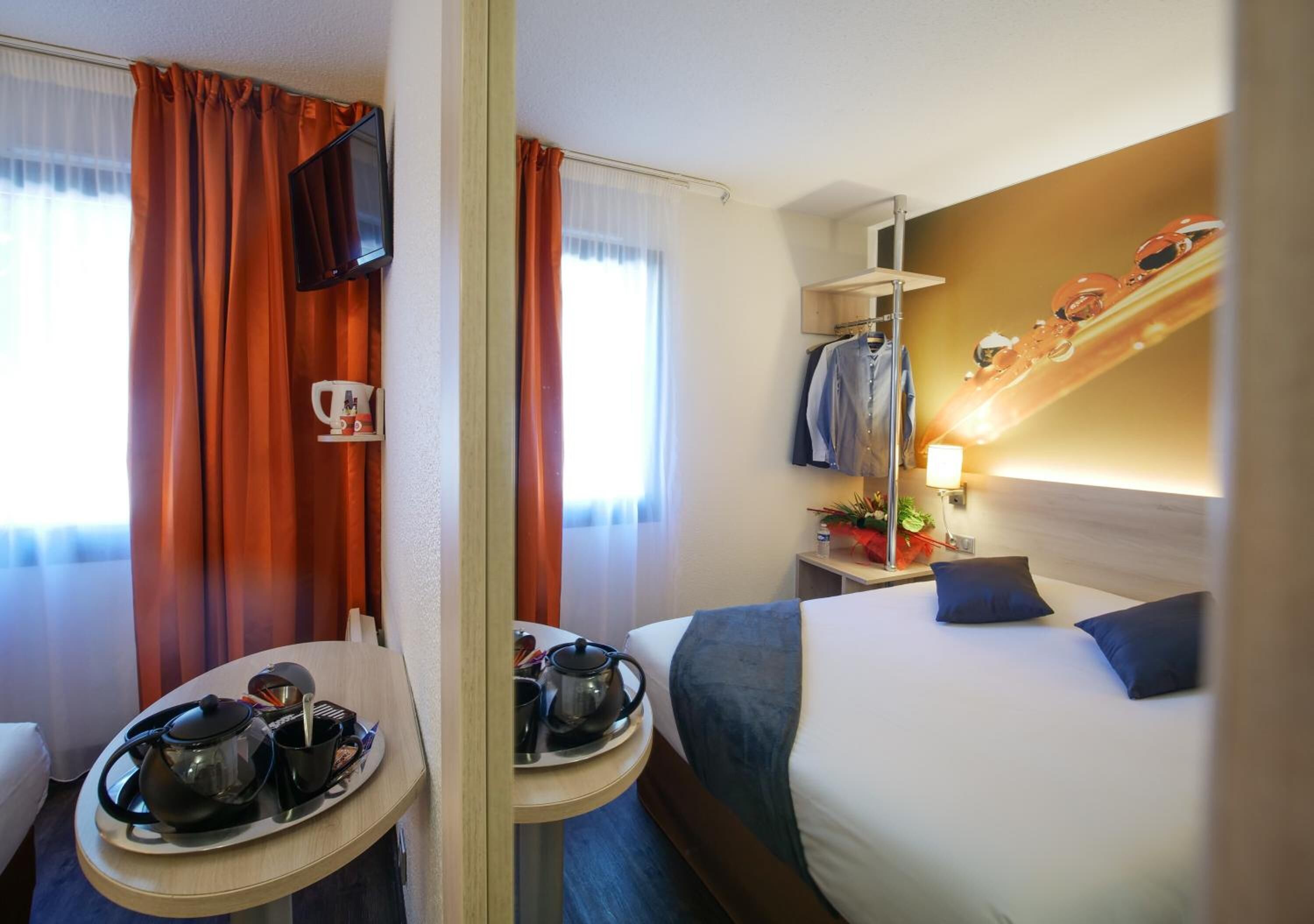 Quarto de hotel com cama, mesa de cabeceira com bandeja de chá e xícaras, televisão na parede e janela com cortinas.
