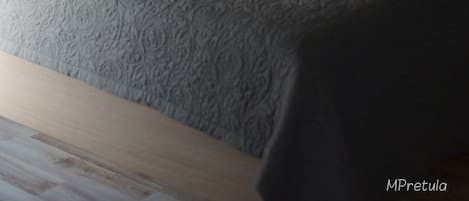 Uma cama bem arrumada com um edredom cinza texturizado, várias almofadas e uma toalha branca dobrada. Uma luminária com cúpula bege está sobre a mesa de cabeceira ao lado da cama.