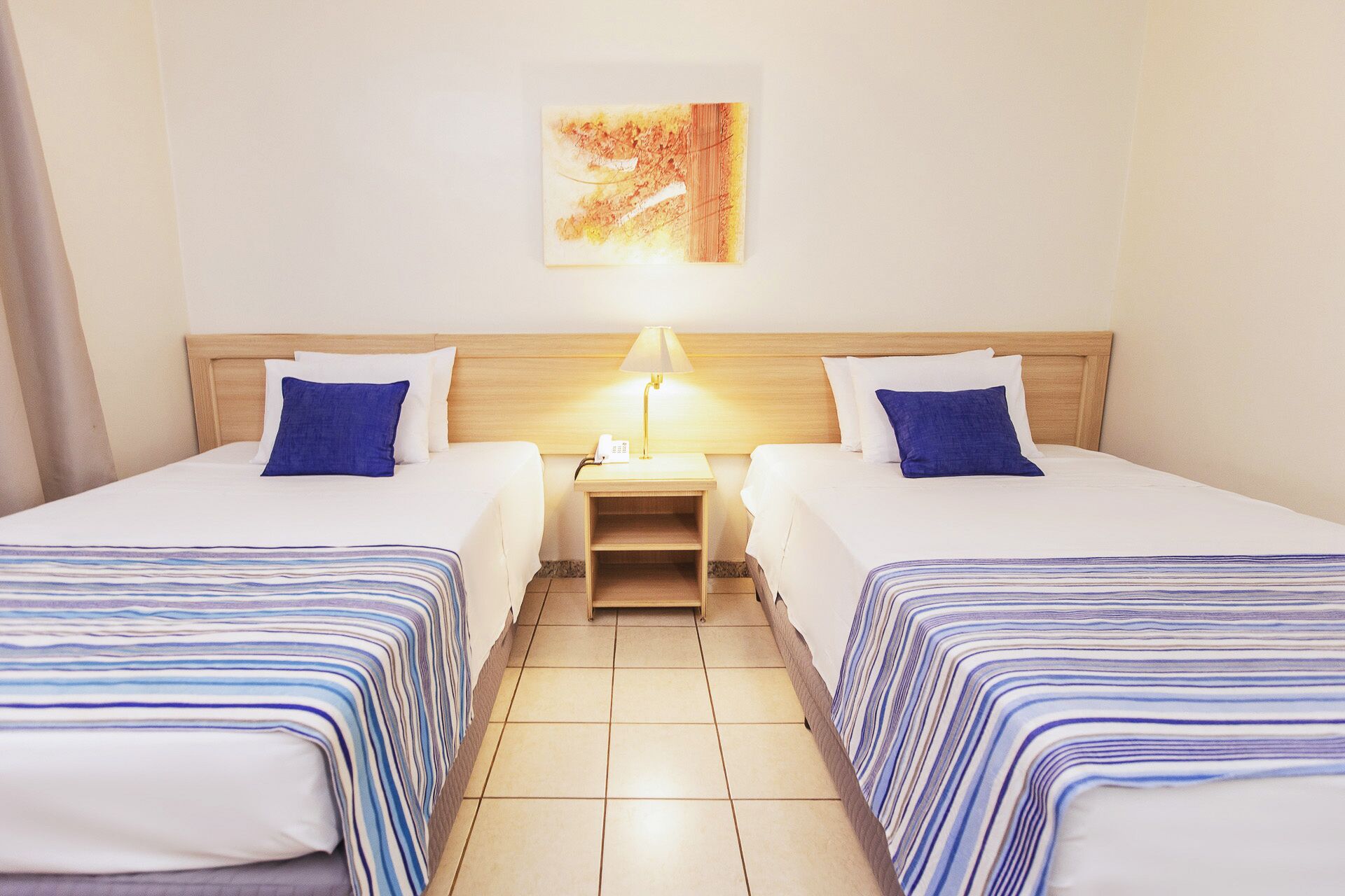 Quarto de hotel com duas camas, cada uma com almofada azul e manta listrada. Há uma mesa de cabeceira com abajur entre as camas.