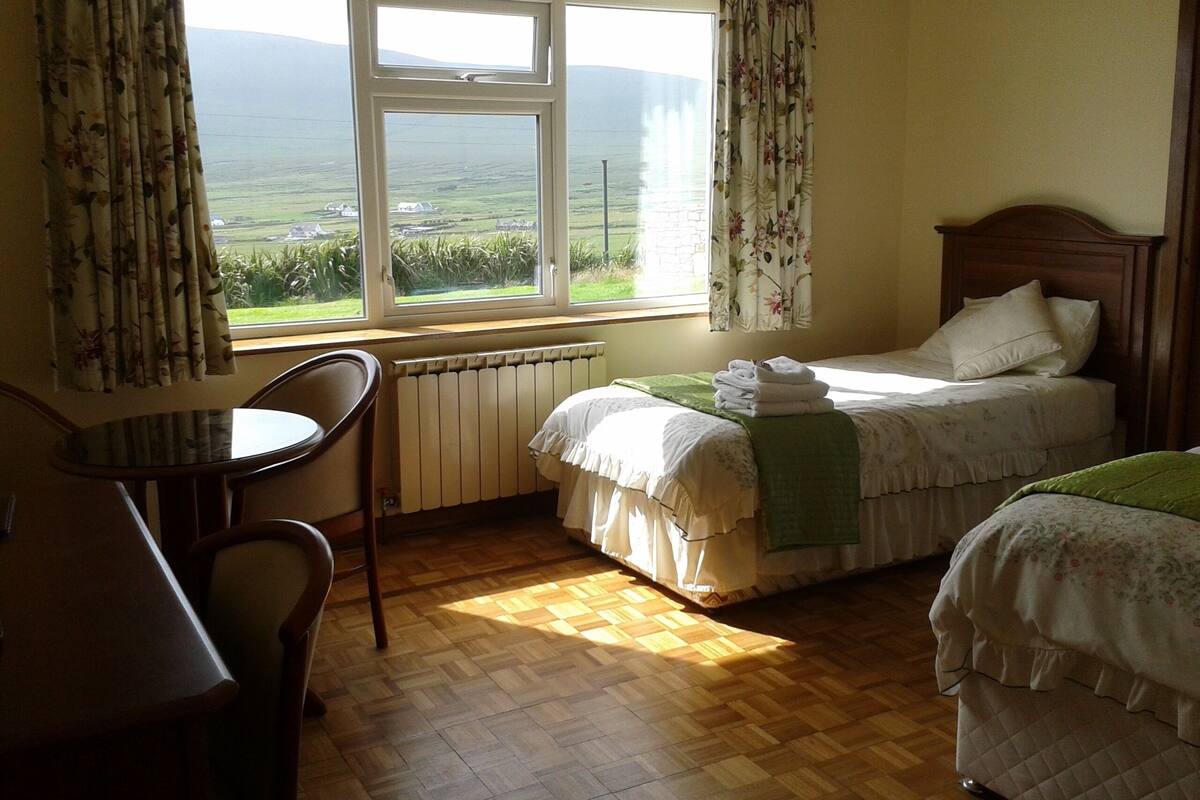 Quarto com duas camas, uma mesa e uma cadeira. Tem uma janela com cortinas, um radiador e vista para o campo.