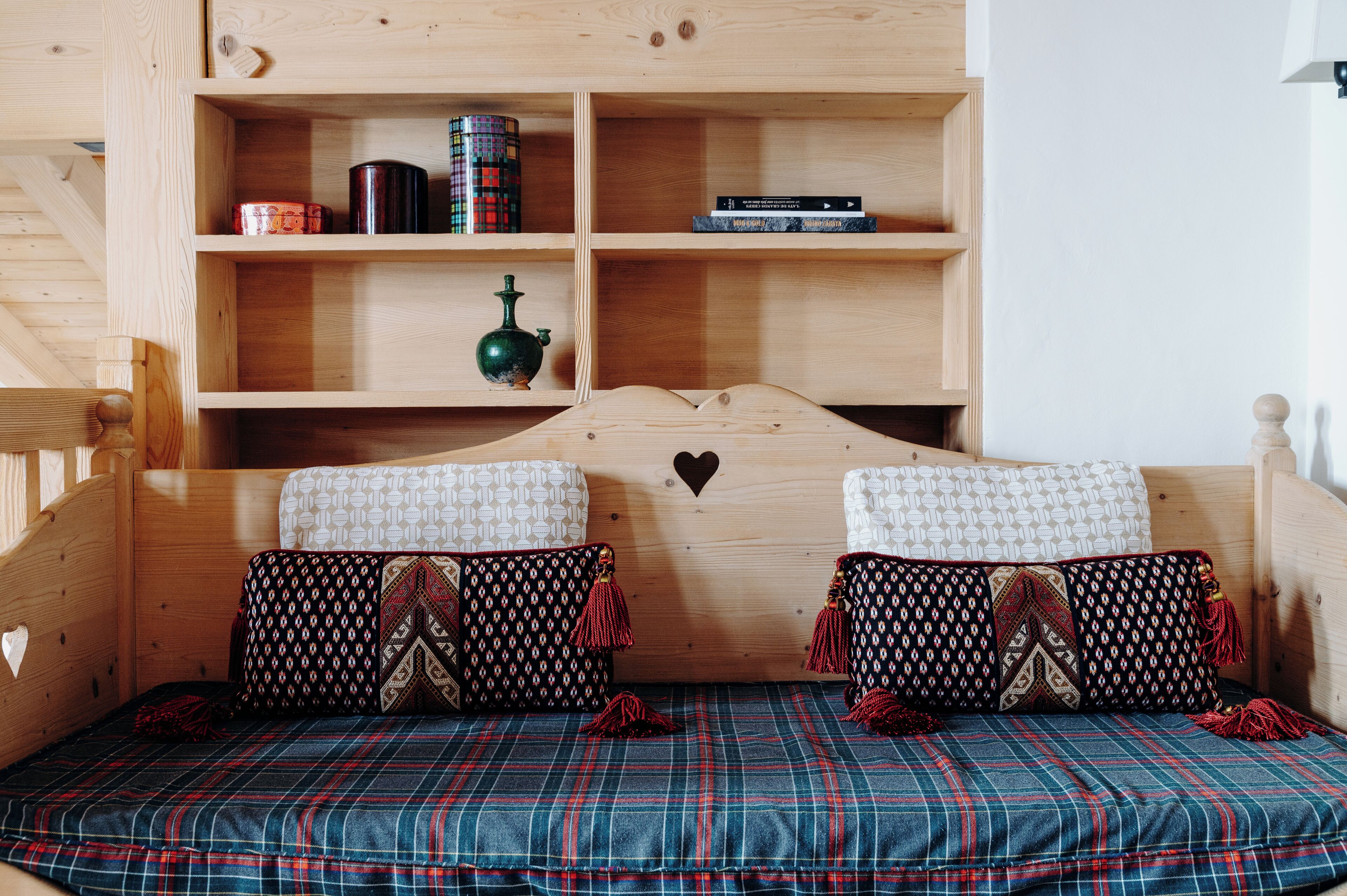 Uma cama beliche de madeira com roupa de cama xadrez, uma estante com livros e objetos decorativos, e um recorte em formato de coração na cabeceira.